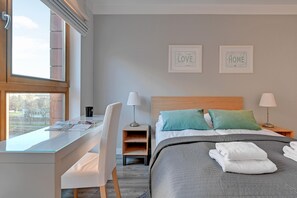 Quarto de hotel com cama, escrivaninha, cadeira, janela e duas quadros na parede.