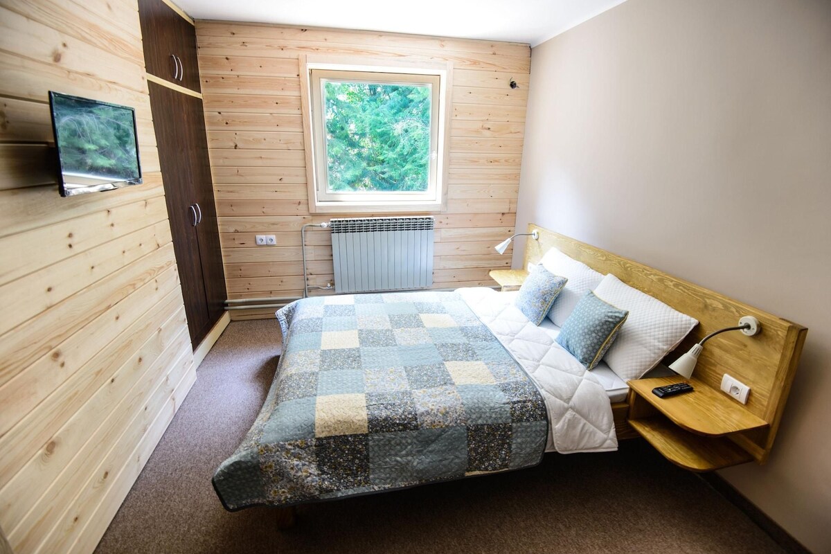 Quarto com cama, mesa de cabeceira, abajur, TV na parede e janela com vista para árvores.