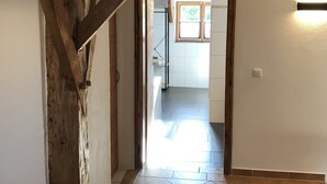 Corredor com vigas de madeira aparentes, que dá para uma cozinha com azulejos brancos e uma janela.