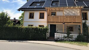 Uma casa de dois andares com painéis solares no telhado, uma varanda de madeira e uma parede de pedra com plantas.