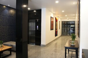 Um lobby moderno com elevador preto, um banco de madeira, uma mesa de vidro com um vaso e obras de arte emolduradas nas paredes.