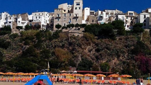Uma cidade costeira com edifícios brancos numa encosta, uma praia com guarda-sóis e uma torre de vigilância.