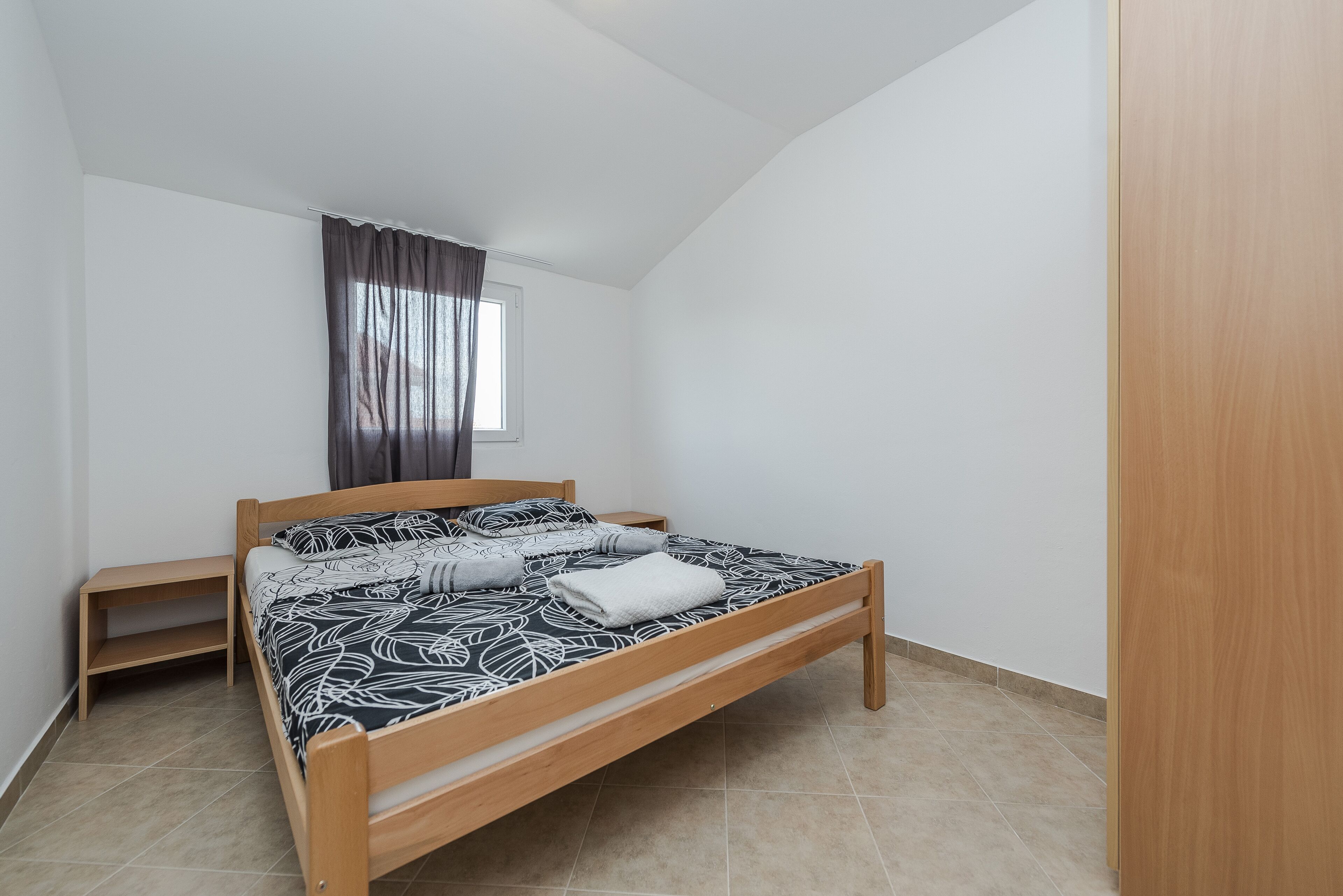 Quarto com cama de madeira, mesa de cabeceira, janela com cortinas e porta.