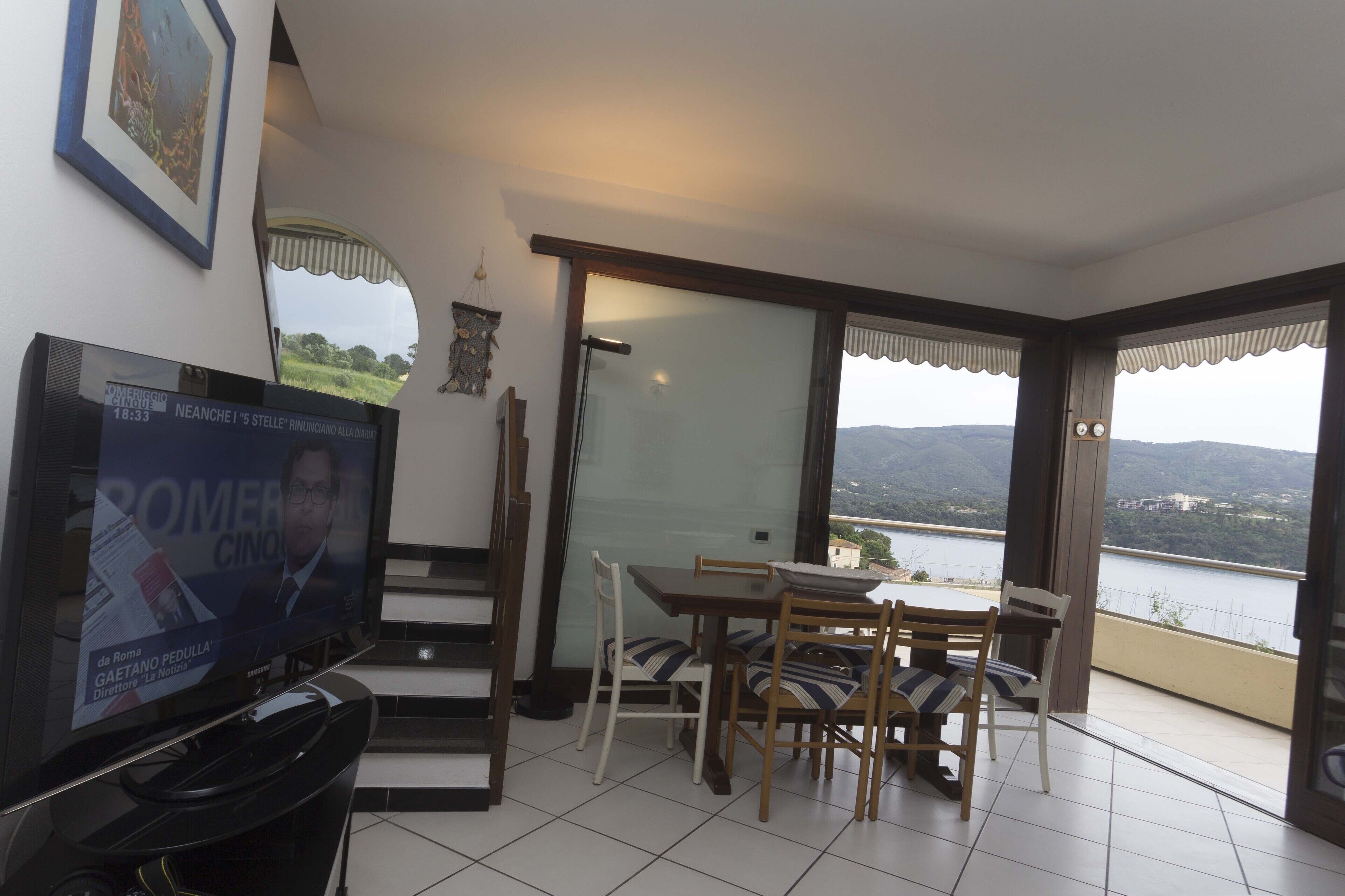 Uma sala de estar moderna com televisão, mesa de jantar, cadeiras e uma varanda com vista panorâmica.