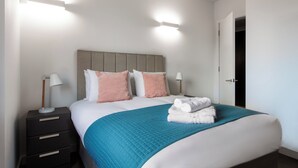 Cama bem arrumada com um edredom verde-água, duas luminárias de cabeceira e uma mesinha de cabeceira com gavetas.