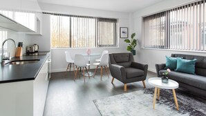 Cozinha moderna com ilha central, sala de jantar com mesa de vidro e cadeiras brancas, sofá cinza com almofadas azul-turquesa e janelas amplas com persianas.