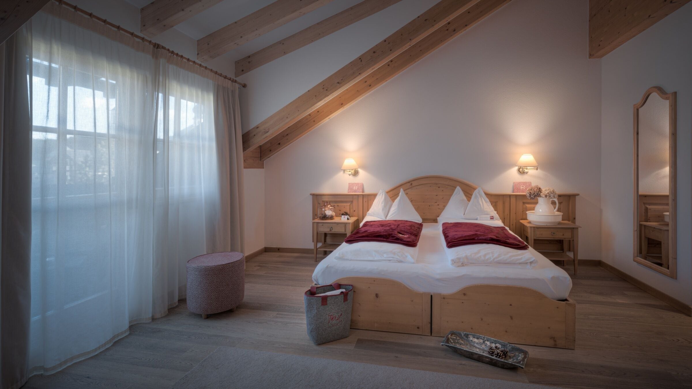 Quarto com cama de madeira, duas mesas de cabeceira, uma janela grande com cortinas leves e teto inclinado com vigas aparentes.