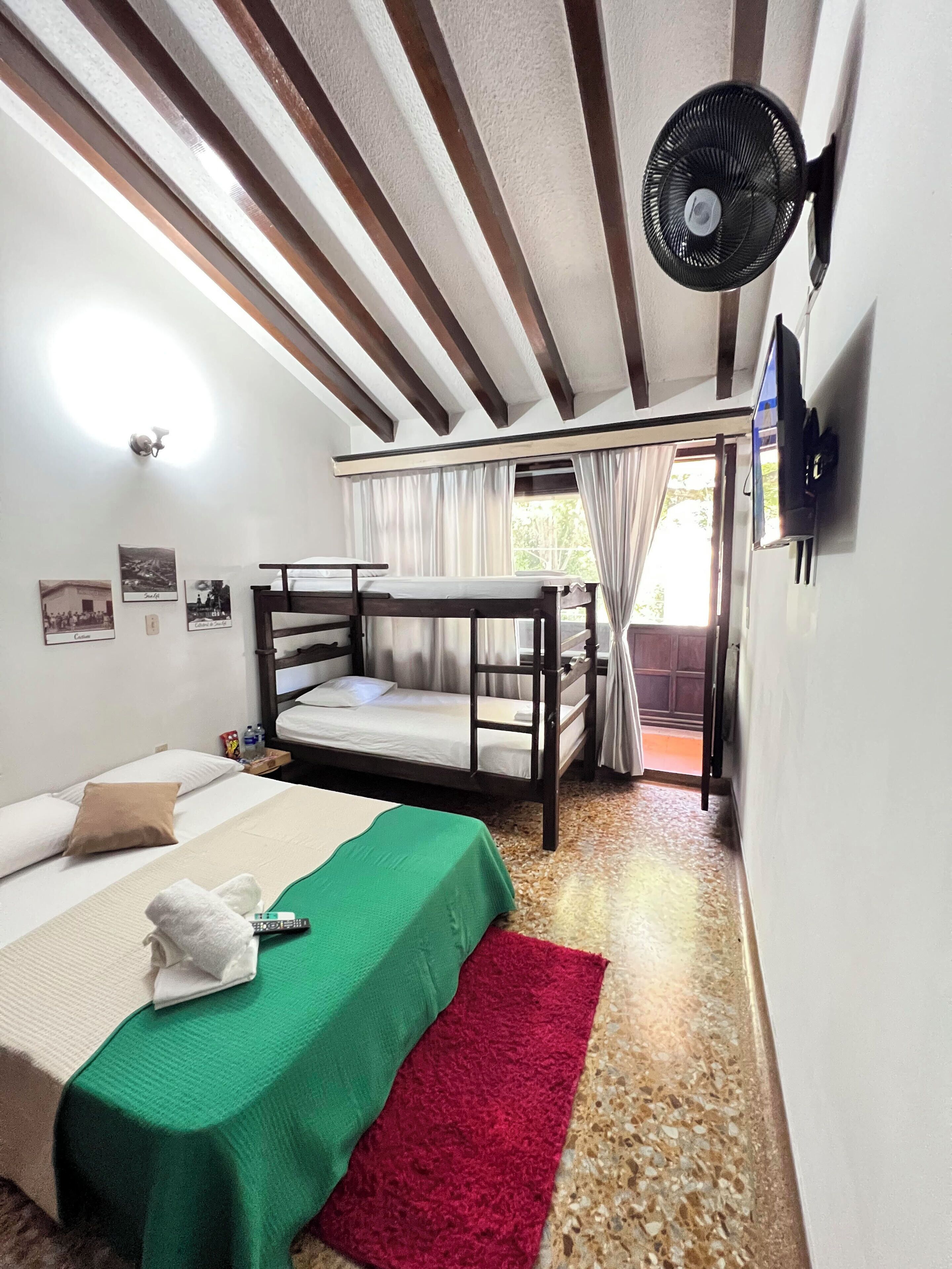 Quarto com cama beliche, uma cama de solteiro, tapete verde e vermelho, ventoinha no teto e uma pequena TV na parede.