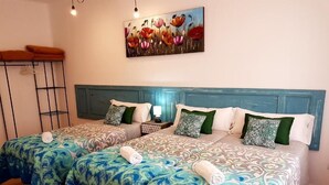 Quarto de hotel com duas camas, cada uma com roupa de cama e almofadas estampadas, um quadro de flores na parede e uma escada encostada na parede.