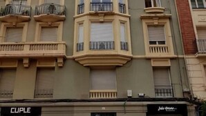 Um edifício de vários andares com uma varanda saliente de esquina, uma loja com a placa 'CUPLE' e um sinal de estacionamento 'P'.