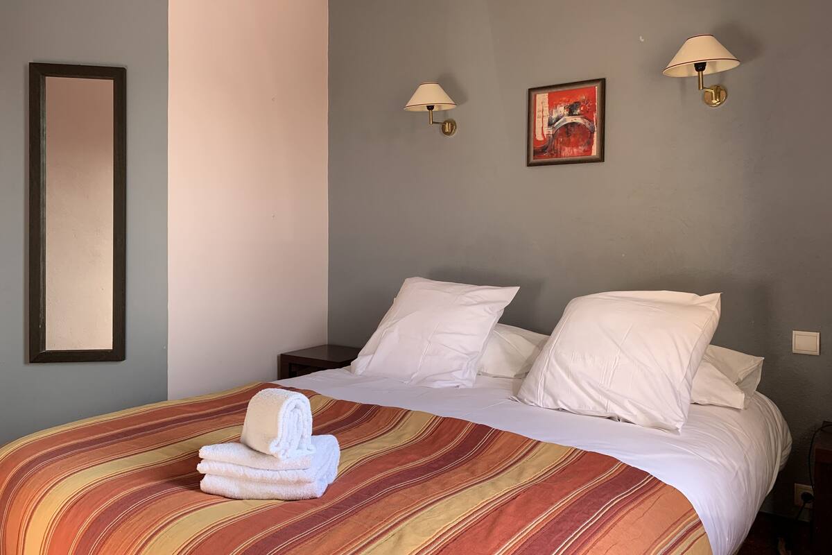 Cama bem arrumada com lençóis brancos, colcha listrada e toalha enrolada no apoio para os pés. Há duas luminárias fixadas na parede e um quadro emoldurado acima da cama.