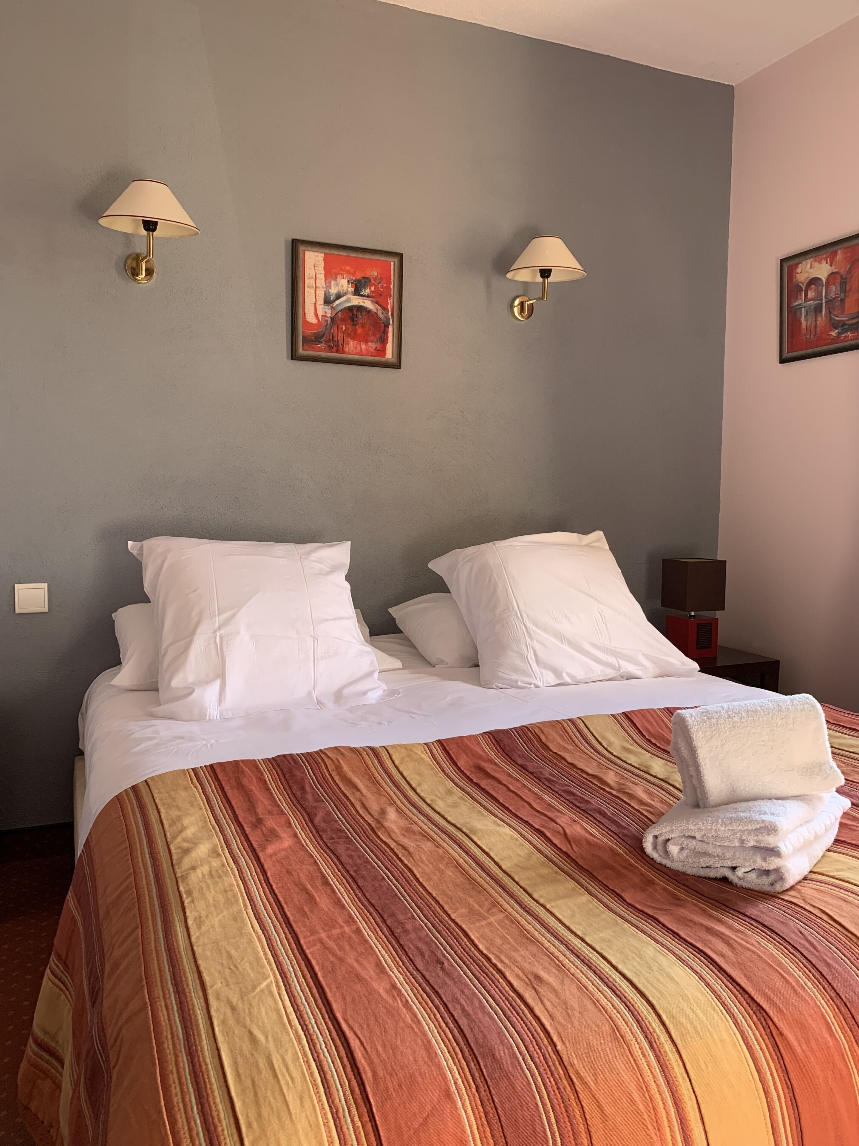 Quarto de hotel com uma cama, duas luminárias fixadas na parede e duas obras de arte emolduradas.