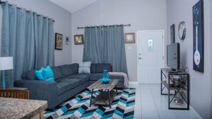 Uma sala de estar moderna com um sofá cinzento, uma mesa de centro e um tapete estampado. Há uma porta com painel de vidro jateado, uma televisão sobre um móvel e um relógio fixado na parede.