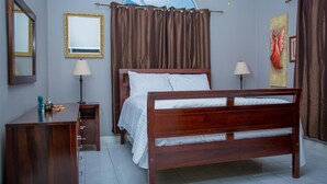 Quarto com cama de madeira, duas mesas de cabeceira, escrivaninha, espelho e janela com cortinas.