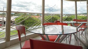 Uma área de estar ao ar livre coberta, com mesas redondas e cadeiras vermelhas, com vista para uma paisagem verde e edifícios ao longe.