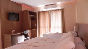 Quarto de hotel com uma cama, duas mesas de cabeceira, uma escrivaninha com cadeira, um guarda-roupa e uma televisão fixada na parede.