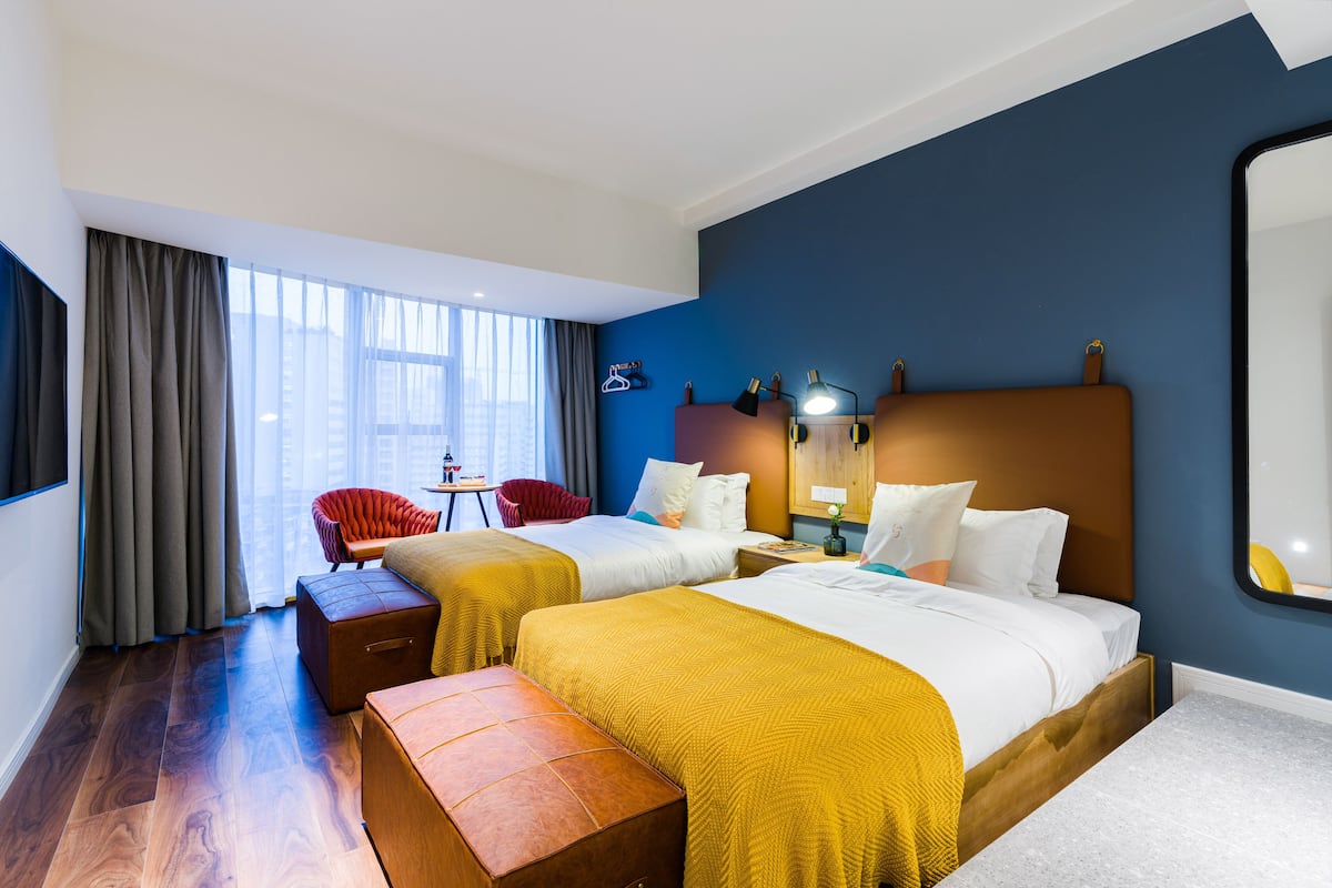 Quarto de hotel com duas camas, uma cadeira vermelha, um banco de madeira e um espelho grande.