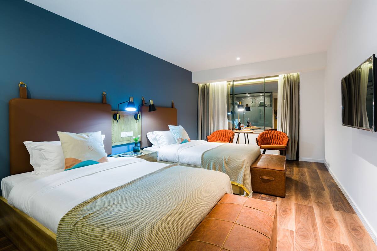 Quarto de hotel com duas camas, uma escrivaninha, uma cadeira e vista para a cidade através de uma porta de vidro.