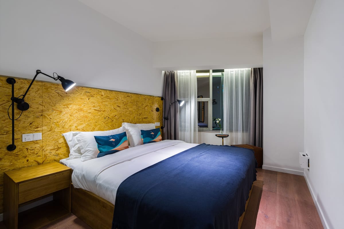 Quarto com cabeceira de madeira, cama com colcha azul, luminária de cabeceira e janela com cortinas.