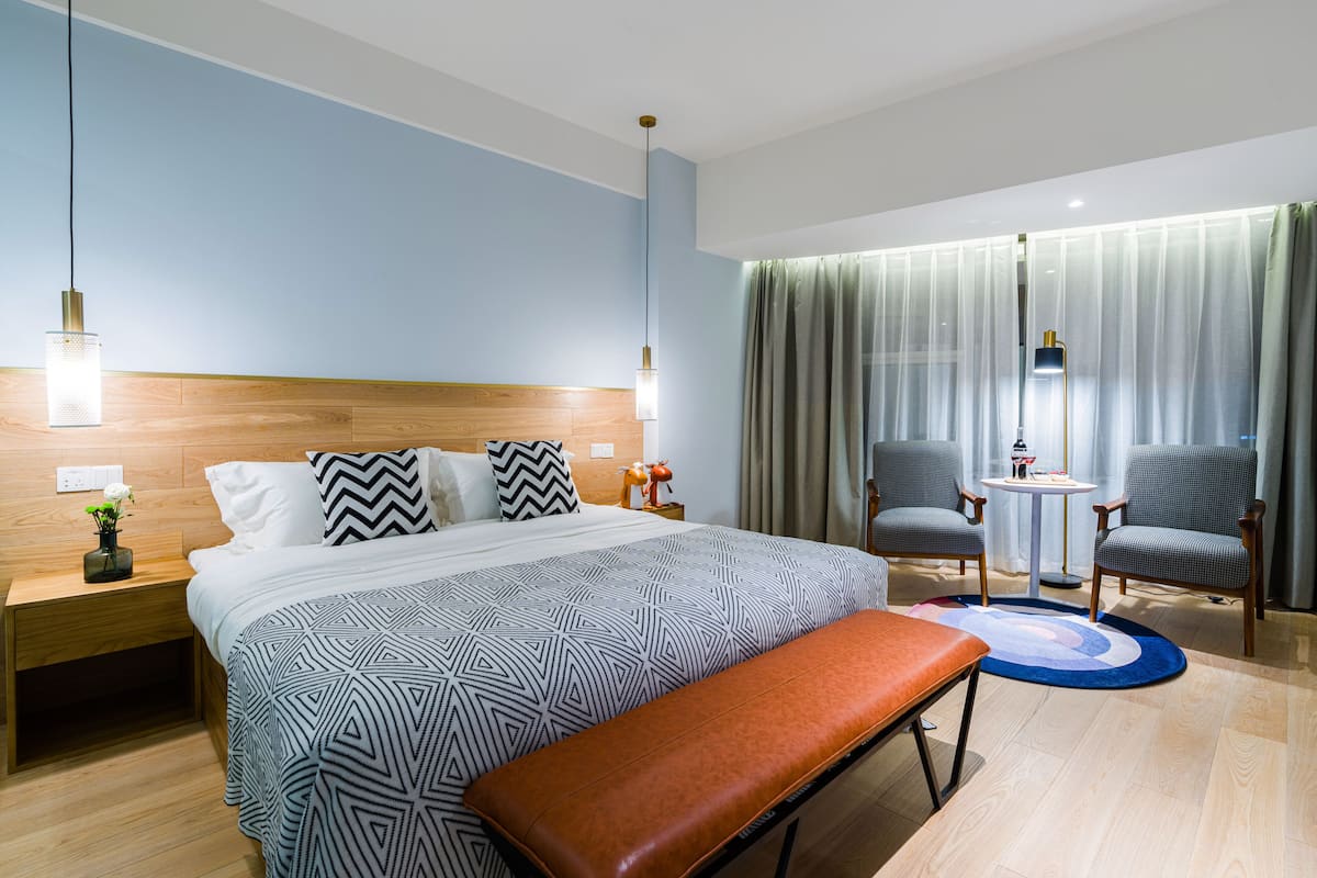 Quarto de hotel moderno com uma cama grande, duas poltronas, uma mesa redonda pequena e um banco.