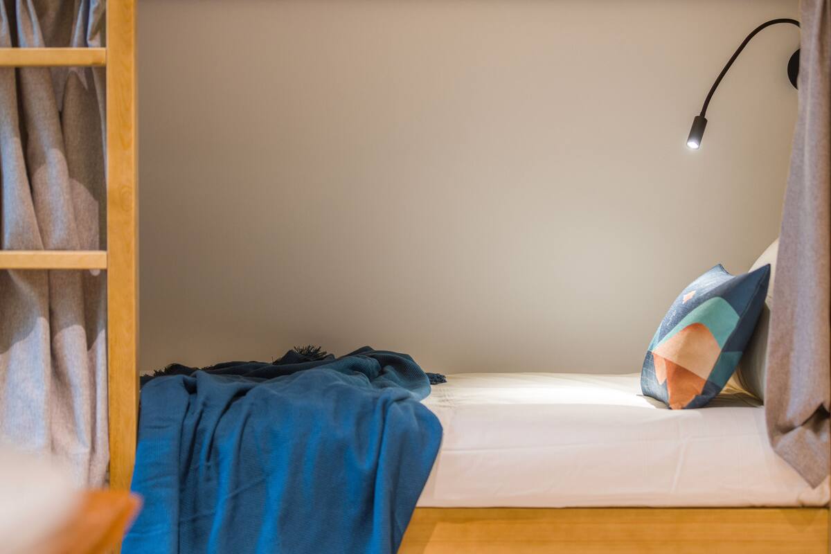 Uma cama de solteiro com um cobertor azul, um travesseiro e uma luminária de cabeceira em um quarto com cabeceira de madeira e parede lisa.