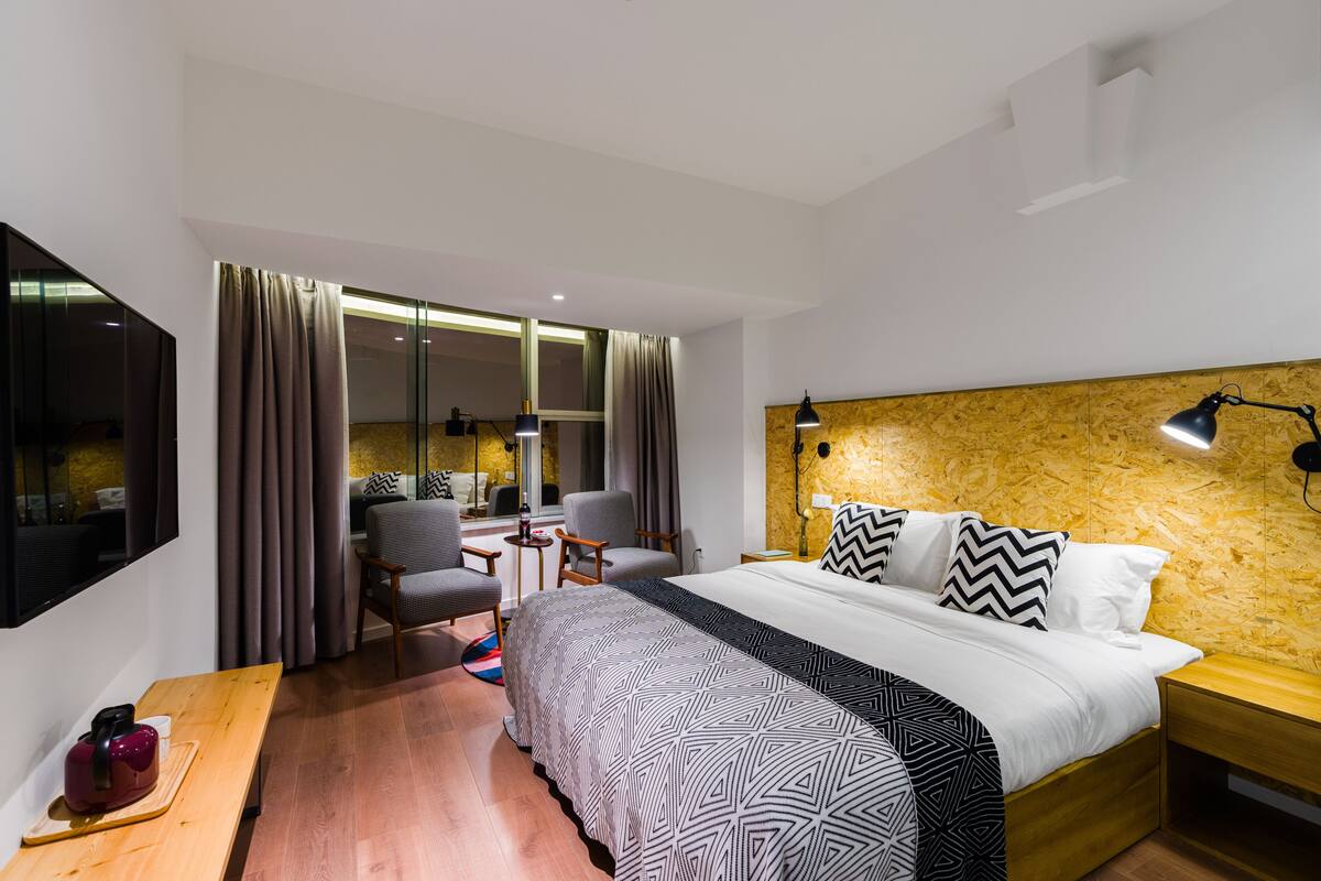 Quarto de hotel moderno com uma cama grande, cabeceira de madeira, escrivaninha com abajur, uma cadeira e vista para a cidade pela janela.