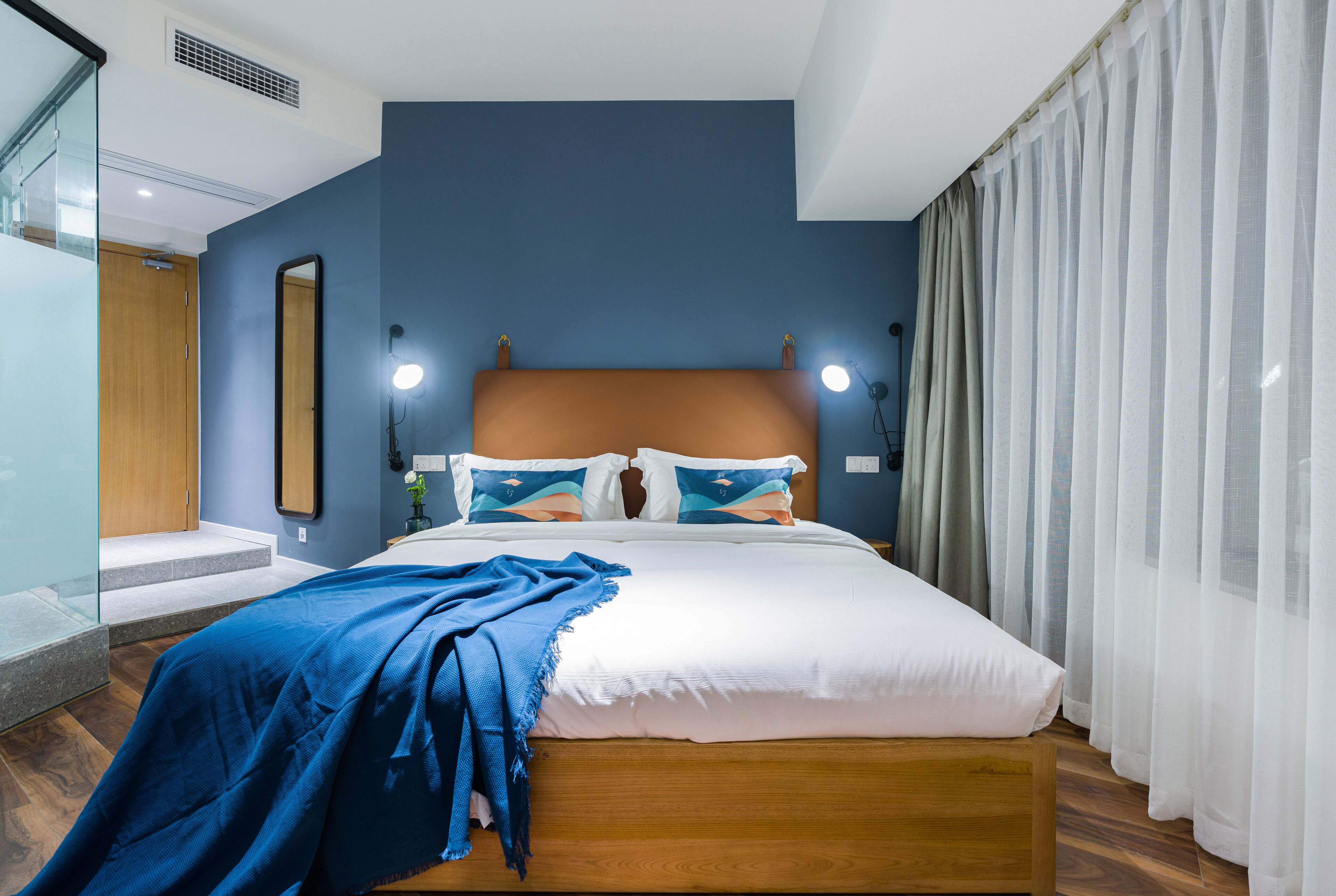 Quarto moderno com cama de madeira, paredes azuis e uma janela grande com cortinas leves.