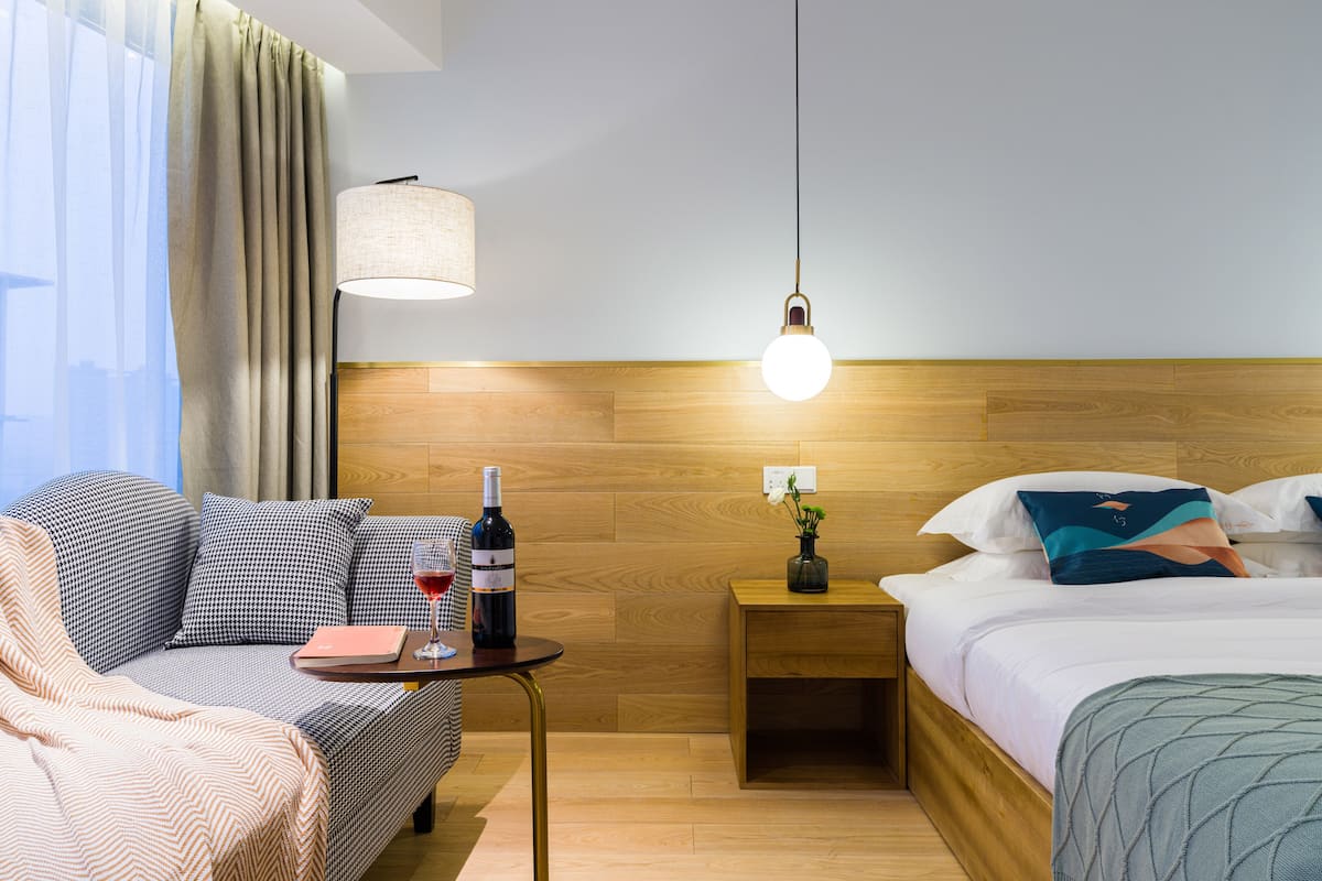 Quarto de hotel com cabeceira de madeira, cama com roupa de cama branca, mesa de cabeceira com abajur, sofá com almofadas estampadas e janela com cortinas.