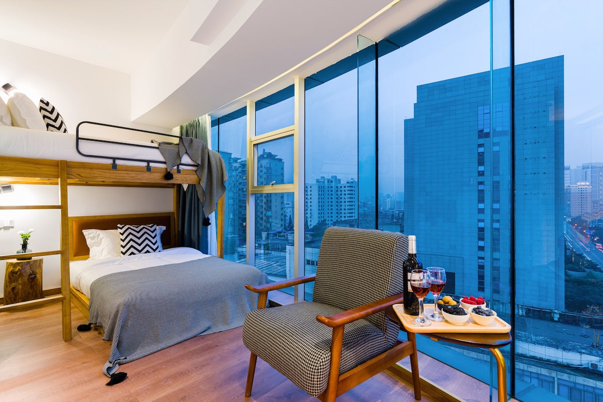 Quarto de hotel moderno com beliche, uma poltrona simples, uma mesa pequena com petiscos e bebidas, e uma janela grande com vista para a cidade.