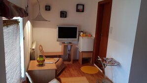 Uma pequena sala de estar com televisão, geladeira, uma mesinha com um vaso e uma mesa de vidro com revistas.
