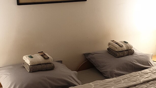 Cama bem arrumada com cobertura acolchoada, duas almofadas e um cobertor dobrado sobre piso de madeira.