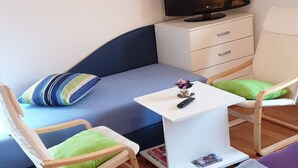 Um quarto moderno e bem iluminado com um sofá, uma cadeira, uma mesinha, uma televisão e um relógio na parede.