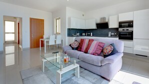 Uma sala de estar moderna com uma mesa de centro de vidro, um sofá cinza com almofadas estampadas, uma área de cozinha com armários brancos e uma área de jantar com mesa e cadeiras brancas.