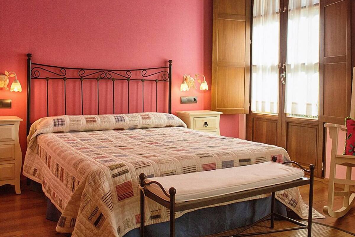 Quarto com estrutura de cama em metal, colcha acolchoada, mesa de cabeceira com abajur e criado-mudo de madeira.