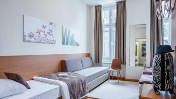 Quarto de hotel moderno com sofá-cama, uma cama individual, uma cadeira e uma mala sobre uma mesinha de madeira.