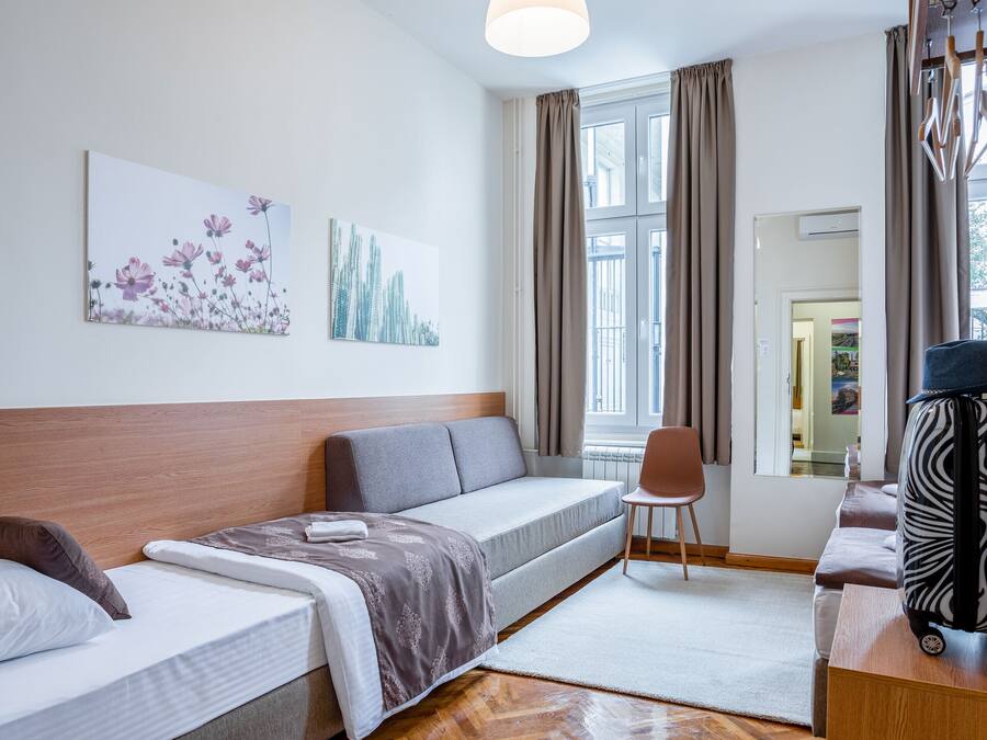 Quarto de hotel moderno com sofá-cama, uma cama individual, uma cadeira e uma mala sobre uma mesinha de madeira.