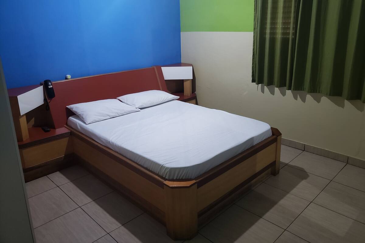 Quarto individual com cama de madeira, roupa de cama branca, mesa de cabeceira com abajur e telefone.