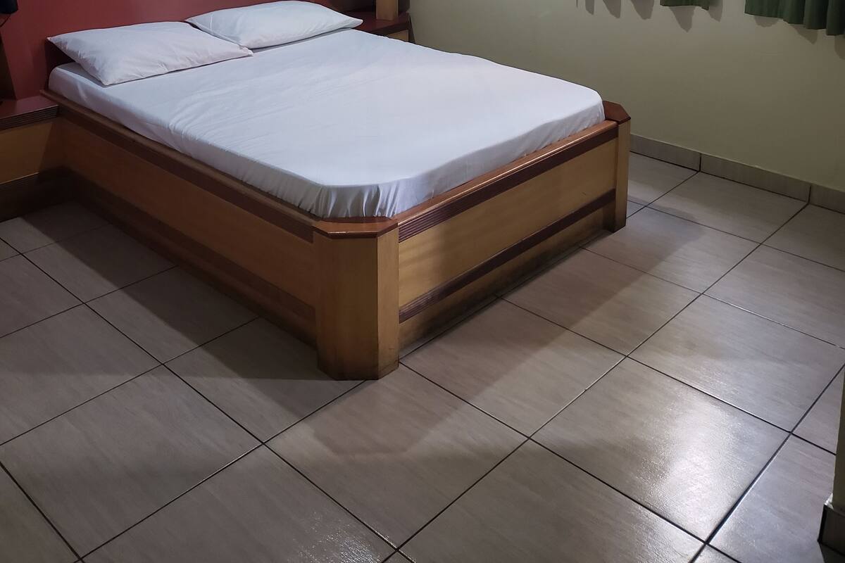 Um quarto com cama, mesa de cabeceira, ventilador de teto e janela com cortinas.