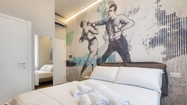 Quarto de hotel com cama, toalhas brancas e um mural de dois homens lutando.
