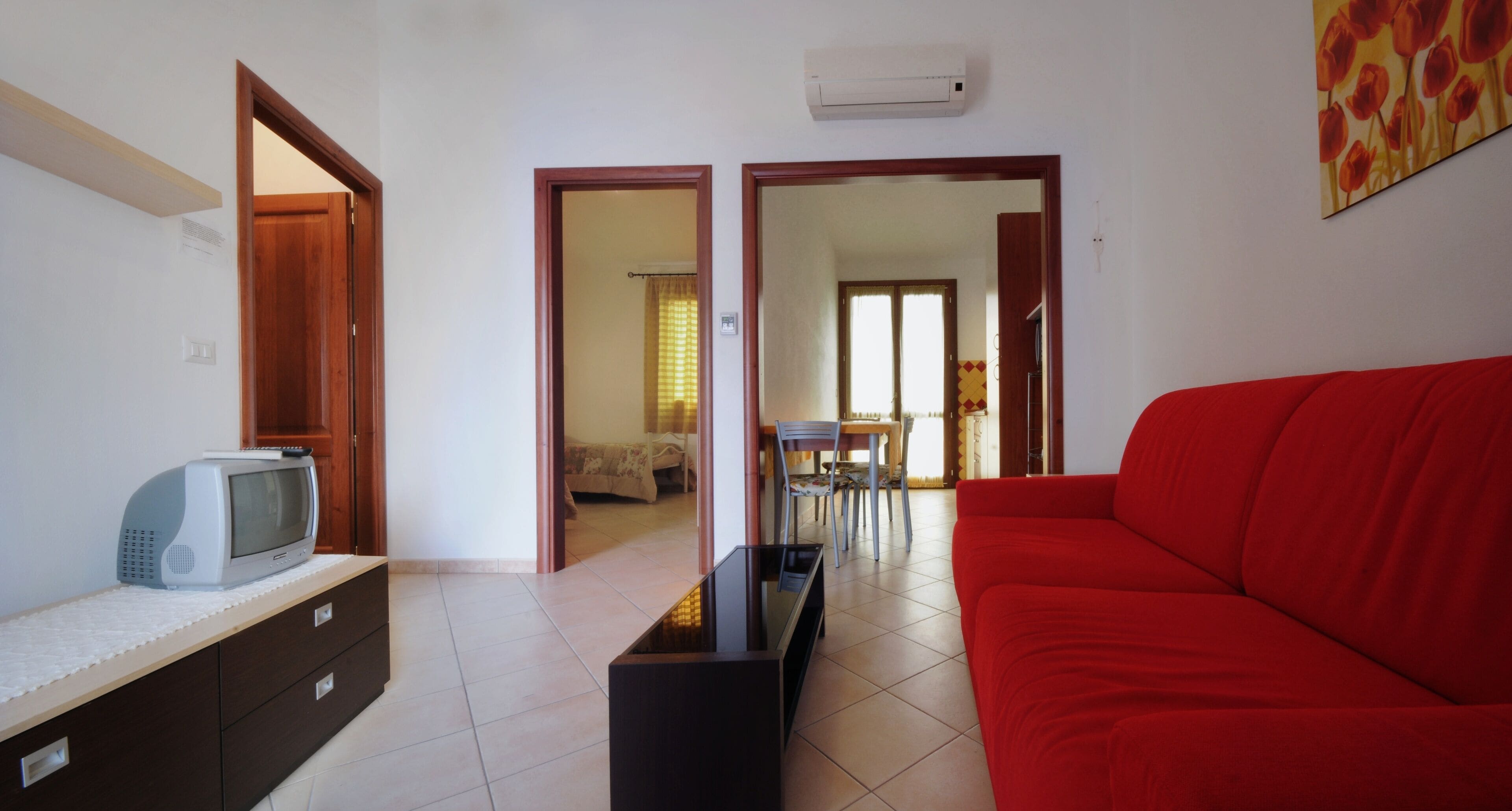 Uma sala de estar com um sofá vermelho, uma televisão sobre um móvel de madeira e uma área de jantar visível através de uma porta aberta.