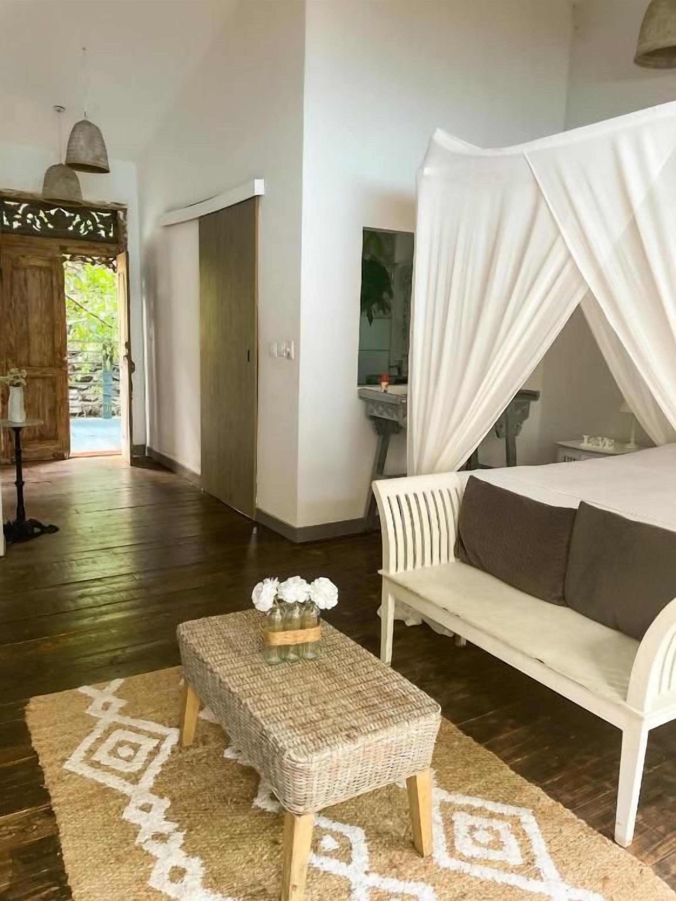 Quarto com sofá branco, mesa de centro de fibra natural, cama com dossel branco e porta de madeira que dá para o exterior.
