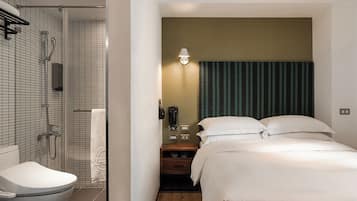 Quarto de hotel com chuveiro, mesa de cabeceira com abajur, cama com almofadas e telefone fixado na parede.