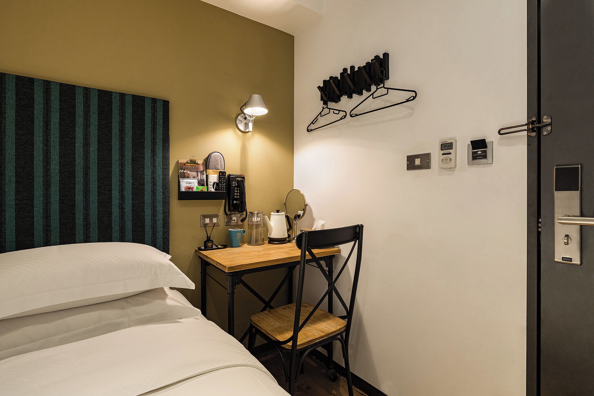 Quarto de hotel com cama, escrivaninha com cadeira, luminária, cafeteira e porta-casacos fixado na parede.