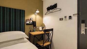 Quarto de hotel com cama, escrivaninha com cadeira, luminária, cafeteira e porta-casacos fixado na parede.
