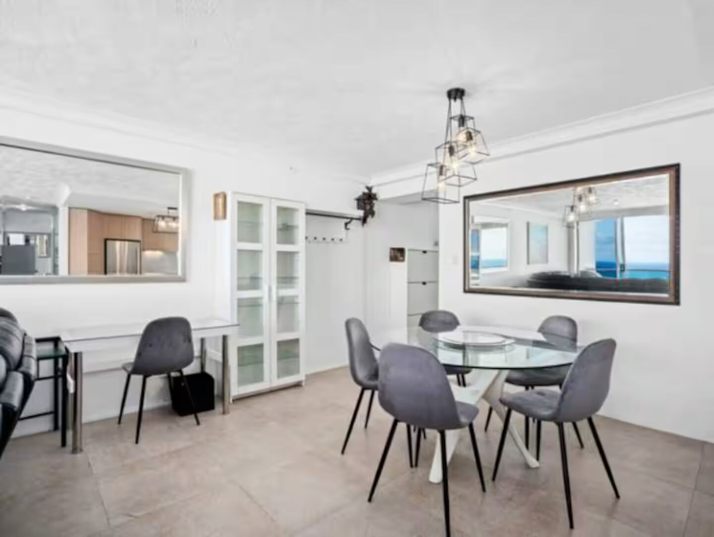 Uma sala de jantar moderna com mesa e cadeiras de vidro, um espelho grande refletindo o ambiente, e vista para a cozinha e sala de estar.