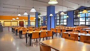 Uma cafeteria com várias mesas e cadeiras, grandes janelas e luminárias pendentes.