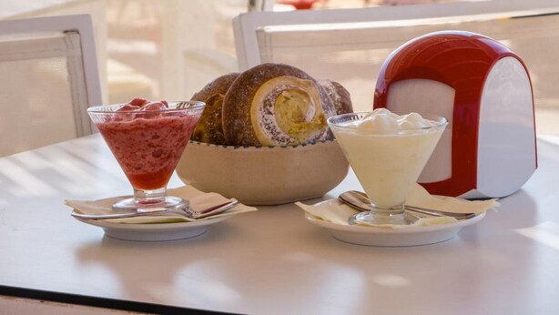 Uma mesa com uma sobremesa, um pão e um recipiente vermelho, com guarda-sóis de praia e veleiros ao fundo.