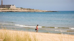 Uma praia com uma pessoa caminhando pela orla, um edifício ao fundo e um céu claro.
