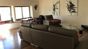 Uma sala de estar espaçosa com sofá de couro, mesa de centro de vidro e área de jantar com mobiliário de madeira. Duas grandes janelas oferecem luz natural e vista para o exterior.