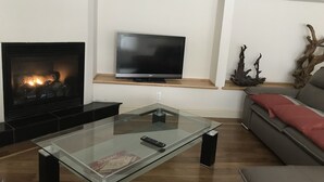 Uma sala de estar com lareira, uma TV de tela plana em uma prateleira de madeira, uma mesa de centro de vidro com controle remoto, e um sofá cinza com almofadas vermelhas.